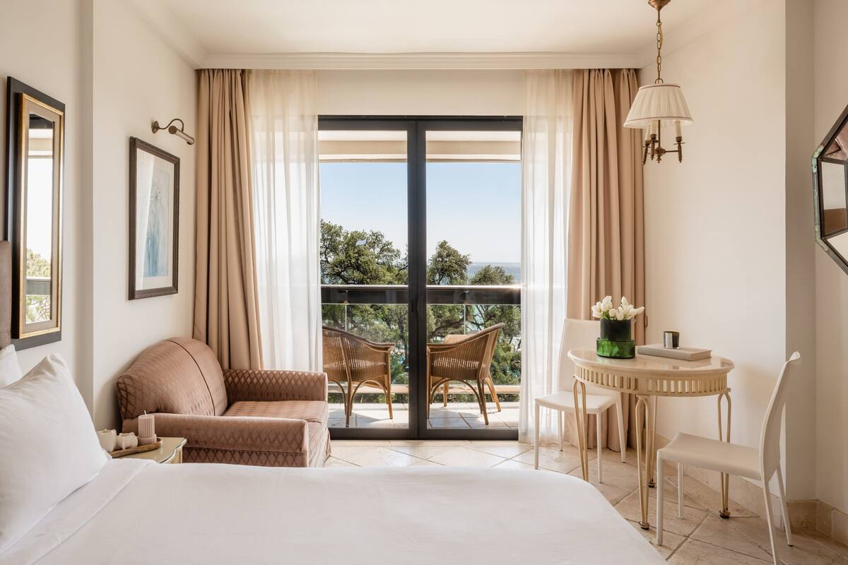 Quarto de hotel com cama, sofá, mesa de jantar com cadeiras e varanda com cadeiras de vime e vista para árvores.
