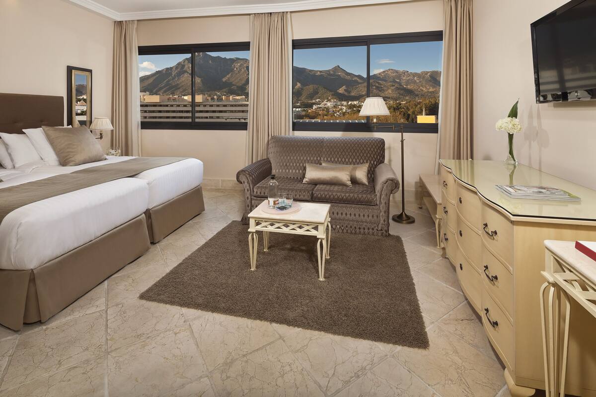 Quarto de hotel com cama, sofá, mesinha, cômoda e televisão.