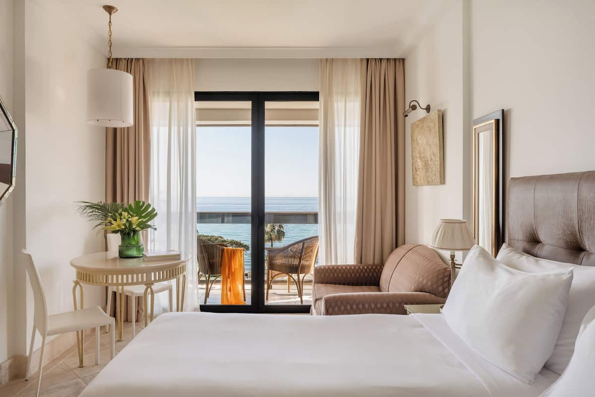 Quarto de hotel com cama, uma pequena mesa com cadeiras, um vaso de planta e vista para o oceano através de uma porta de vidro.
