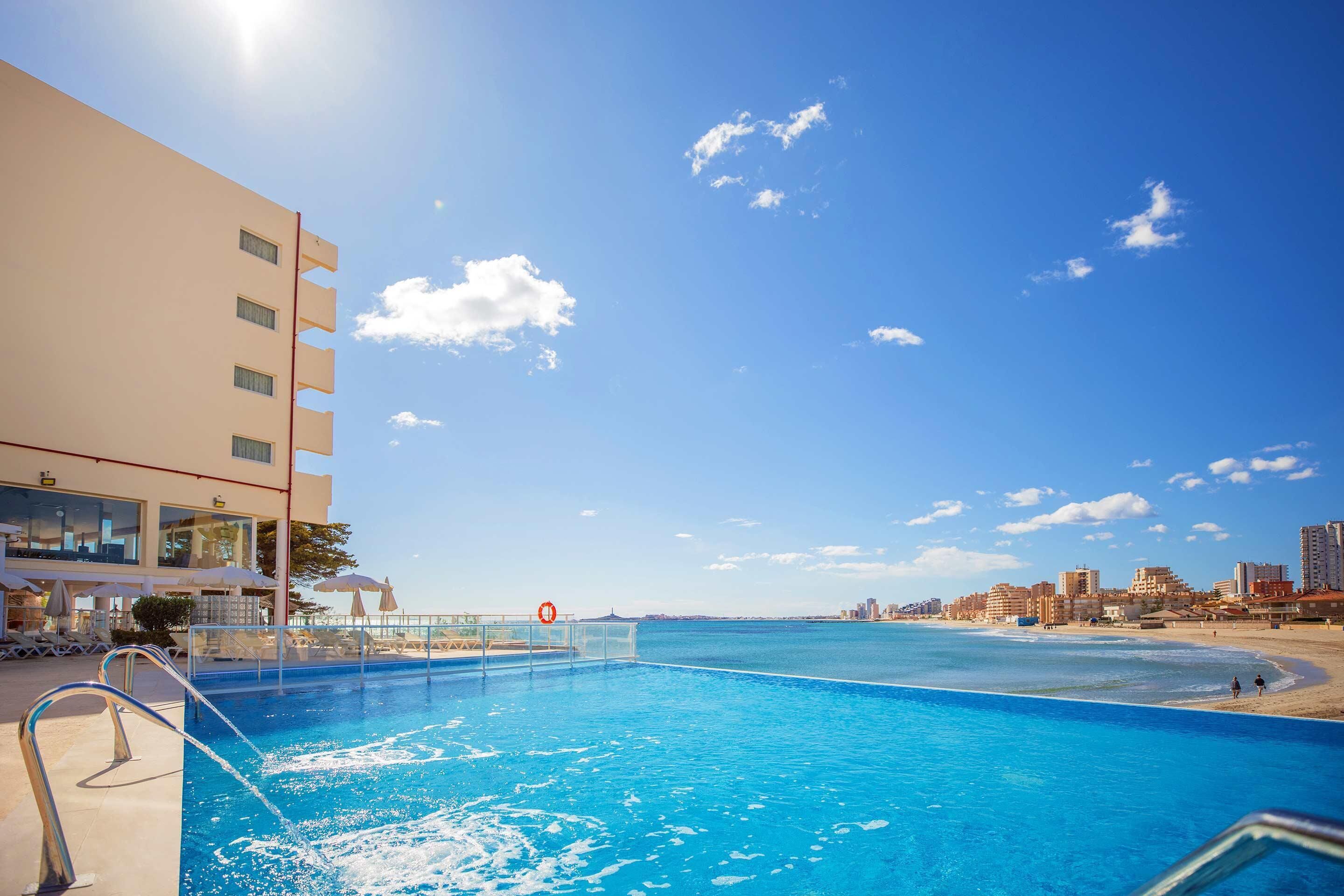 Piscina com água azul cristalina, paisagem urbana ao fundo e uma praia à direita.