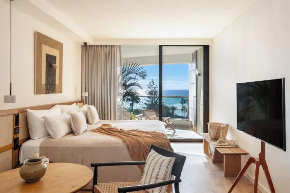 Quarto de hotel com cama, televisão, mesa de jantar e vista para o oceano.