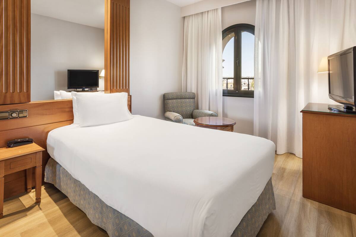 Quarto de hotel com uma cama grande, televisão, escrivaninha com telefone, cadeira e uma pequena mesa.