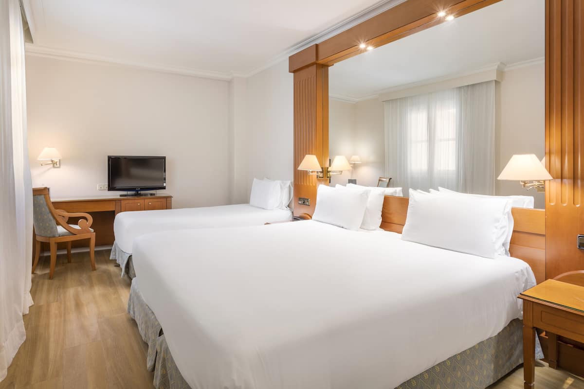 Quarto de hotel com duas camas, uma escrivaninha, uma televisão e duas luminárias.