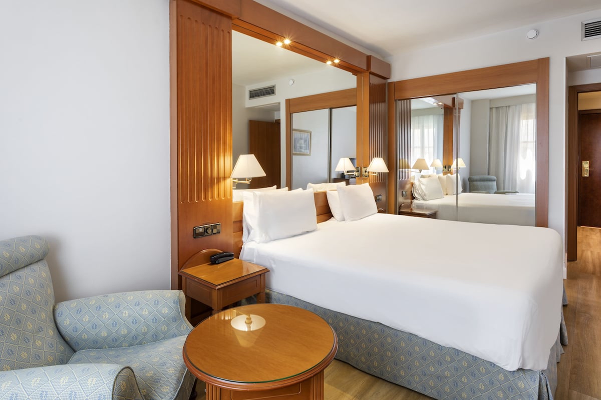 Quarto de hotel com uma cama grande, área de estar com uma poltrona e uma mesinha, e um espelho grande que reflete o quarto.