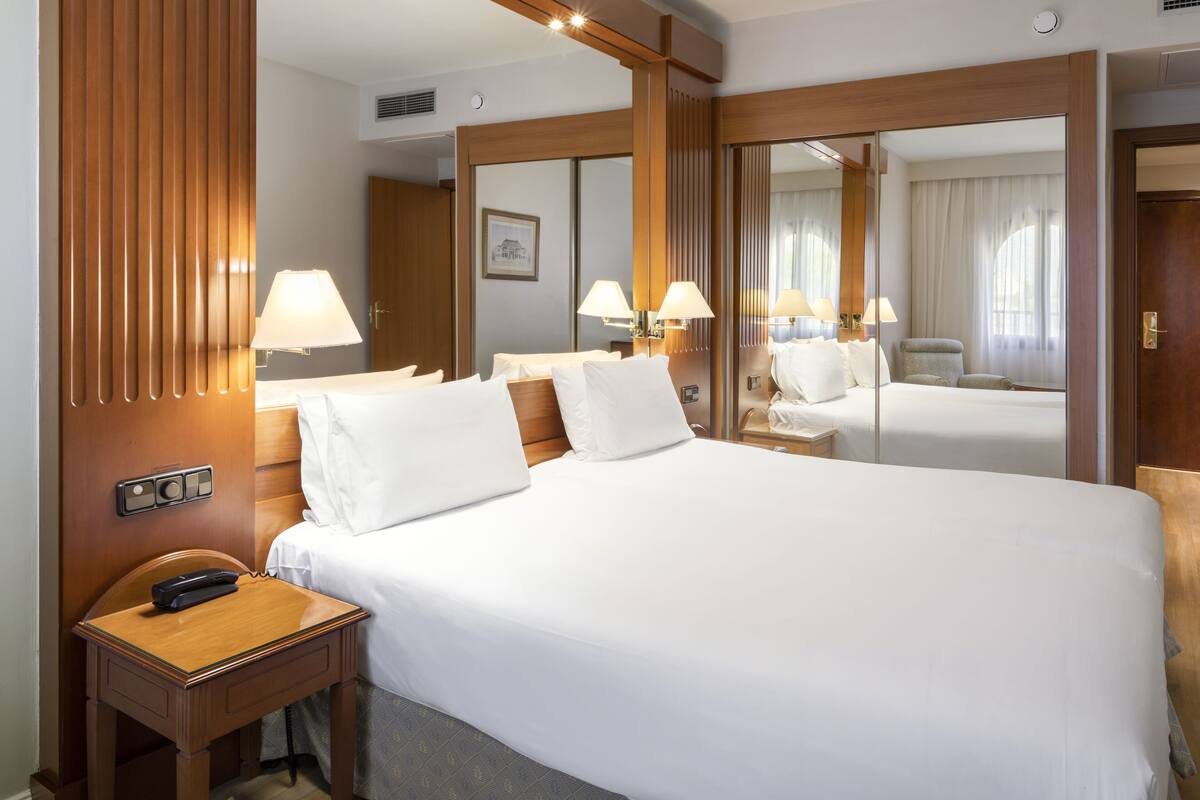 Quarto de hotel com uma cama grande, abajures, mesa de cabeceira de madeira, telefone e um espelho refletindo o aposento.