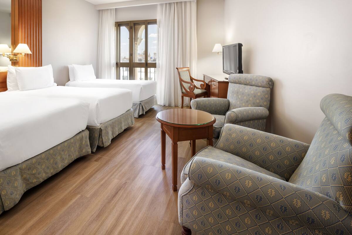Quarto de hotel com duas camas, um sofá, uma poltrona, uma mesinha e uma televisão.