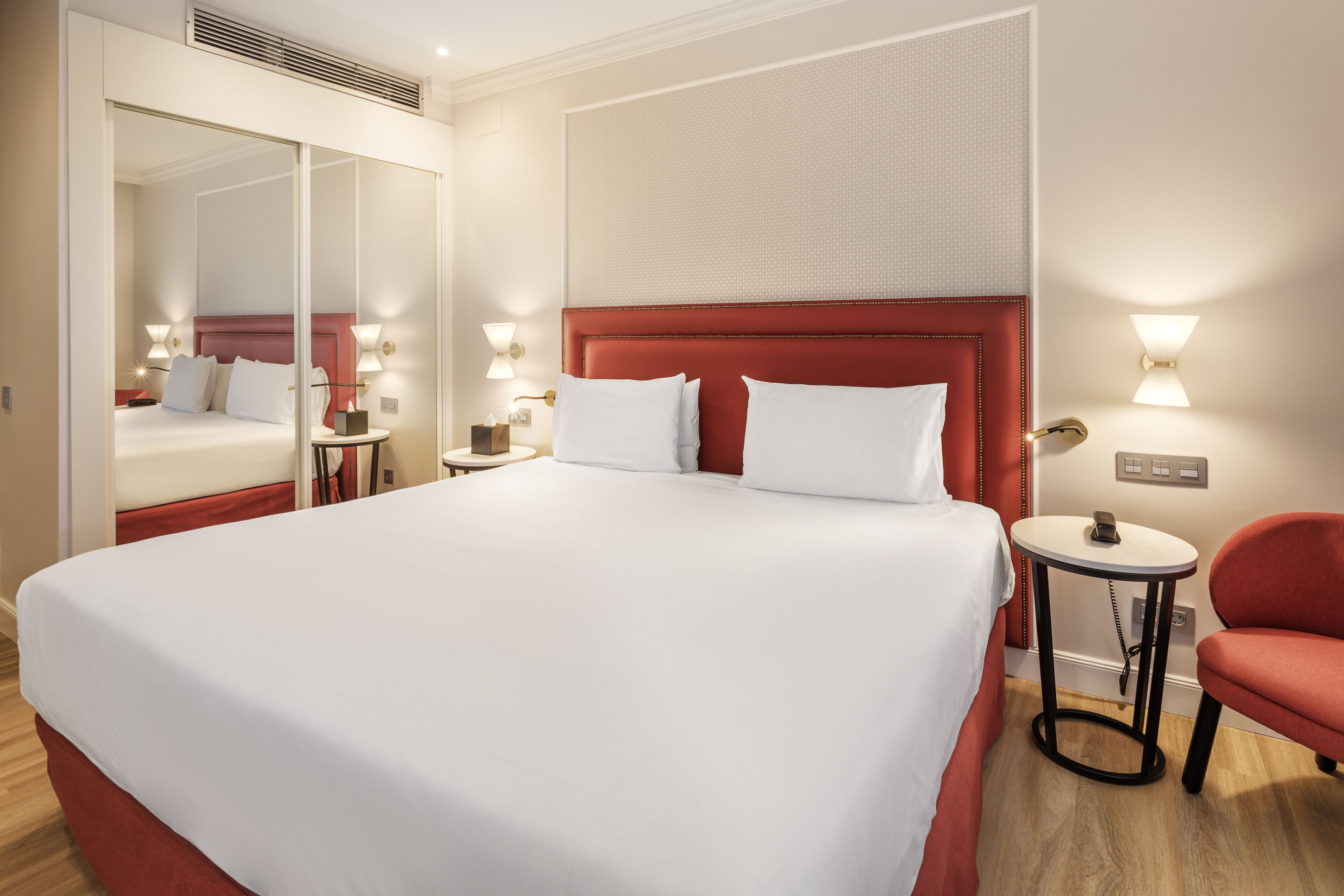 Quarto de hotel com uma cama grande, cabeceira vermelha, mesa redonda, cadeira vermelha e um espelho.