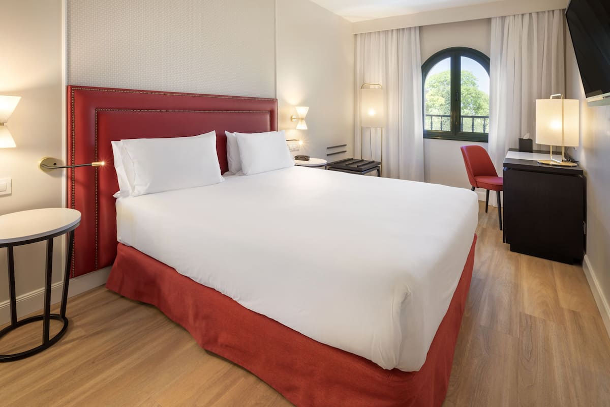 Quarto de hotel com uma cama grande, uma escrivaninha, uma cadeira e uma janela com cortinas.