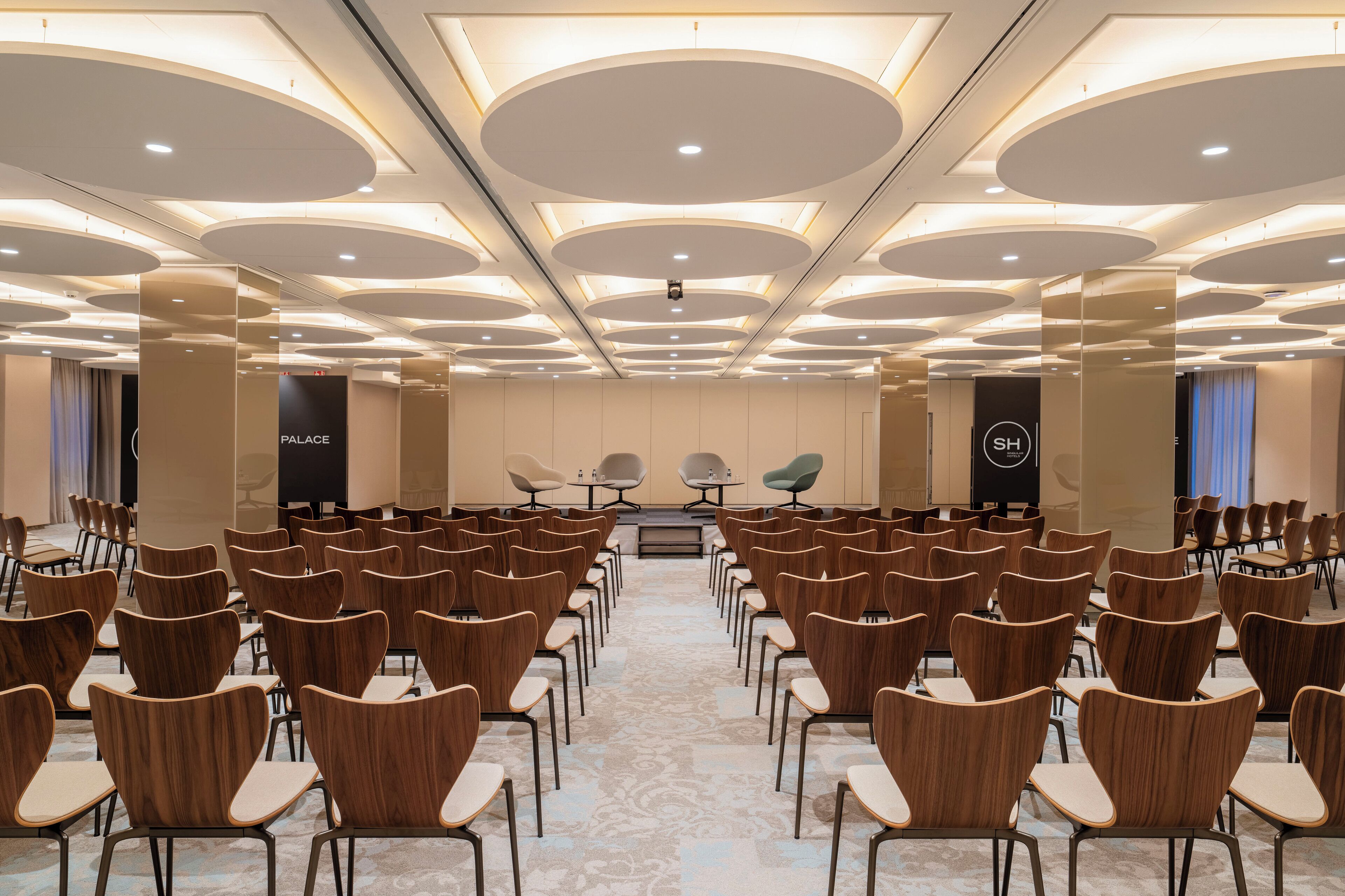 Uma sala de conferências com fileiras de cadeiras de madeira, um palco e um design moderno no teto.