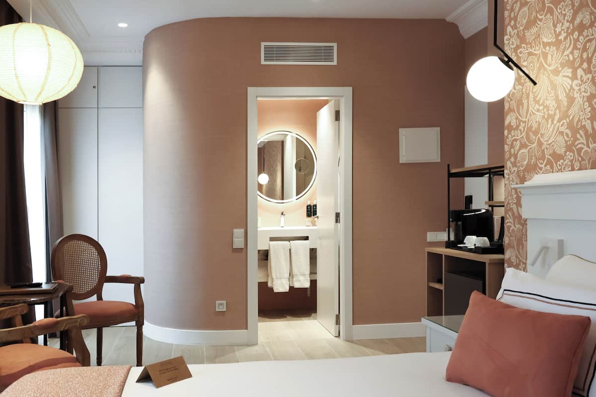 Quarto de hotel com cama, uma cadeira, escrivaninha, abajur, mesa de cabeceira, cafeteira e banheiro visível através de uma porta aberta.