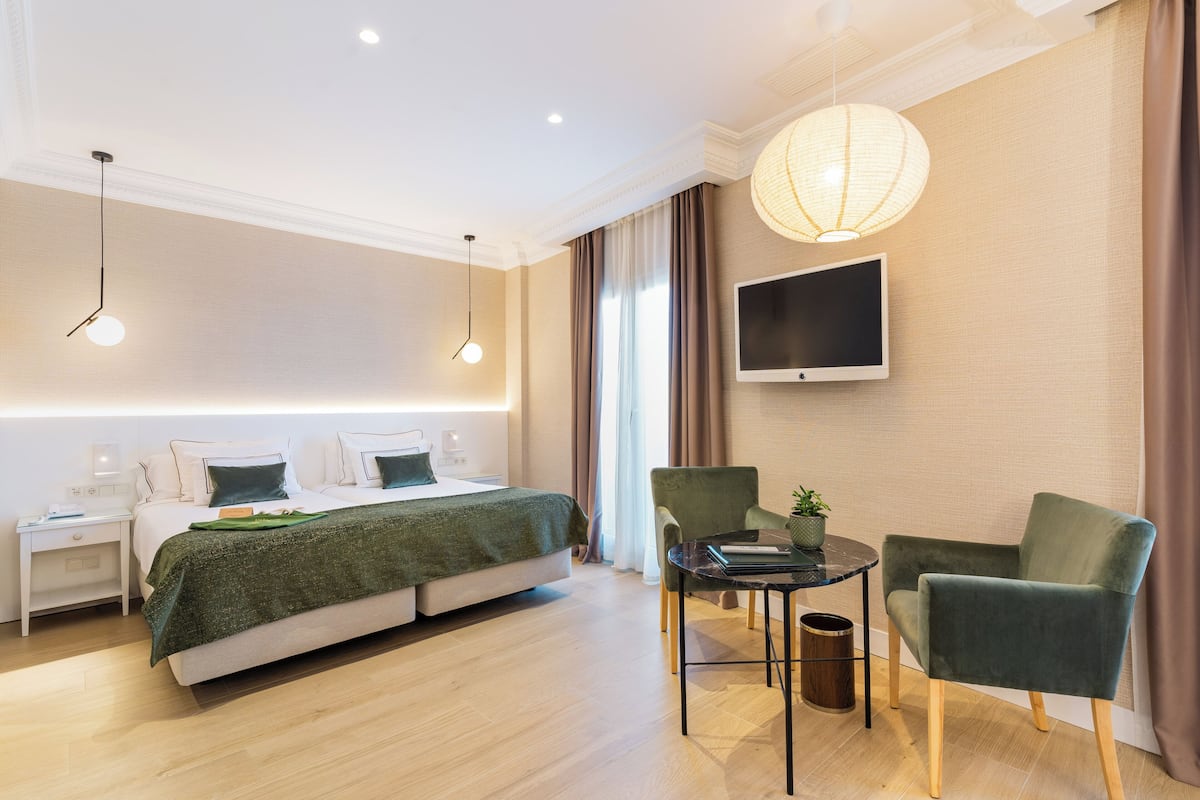 Quarto de hotel moderno com cama, mesas de cabeceira, televisão de tela plana, uma pequena mesa com cadeira e uma janela grande com cortinas.