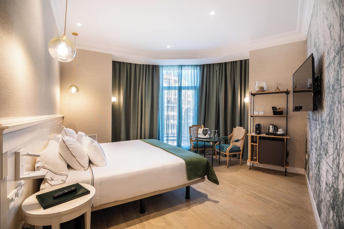 Quarto de hotel com cama, mesa de cabeceira, abajur, cadeira, escrivaninha e televisão na parede.