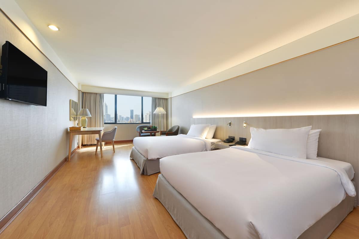 Quarto de hotel com duas camas, uma escrivaninha com cadeira, televisão na parede e uma janela grande com vista para a cidade.