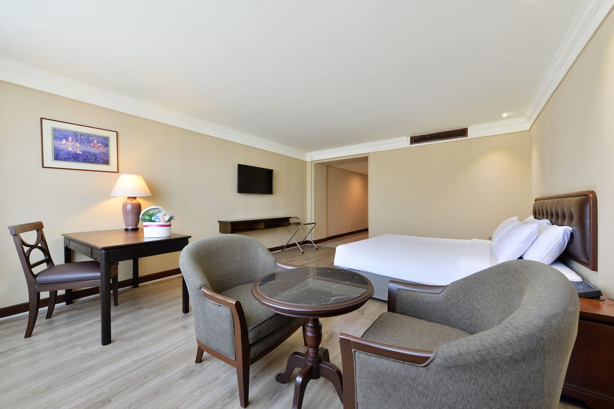 Quarto de hotel com uma cama, duas poltronas, uma mesinha, uma luminária, uma TV e um quadro na parede.