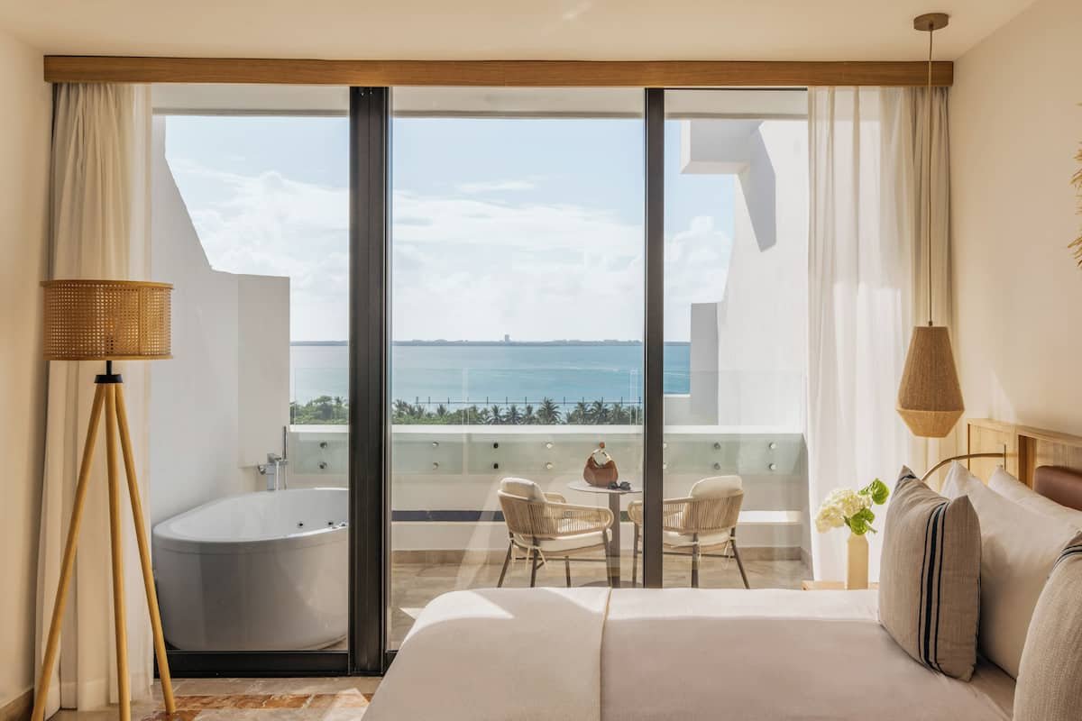 Quarto de hotel moderno com uma cama grande, banheira, varanda com vista para o mar e área de refeições.