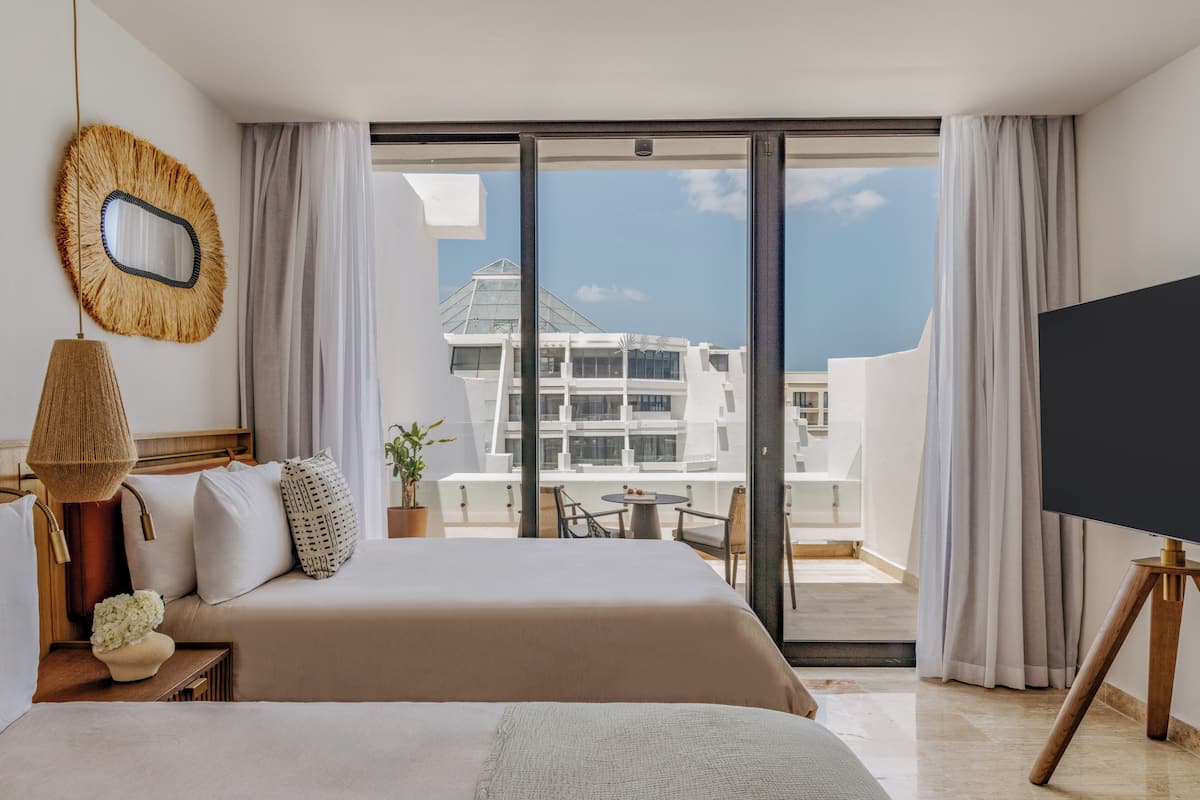 Quarto de hotel com cama, televisão, abajur e vista para um edifício através de uma grande janela.