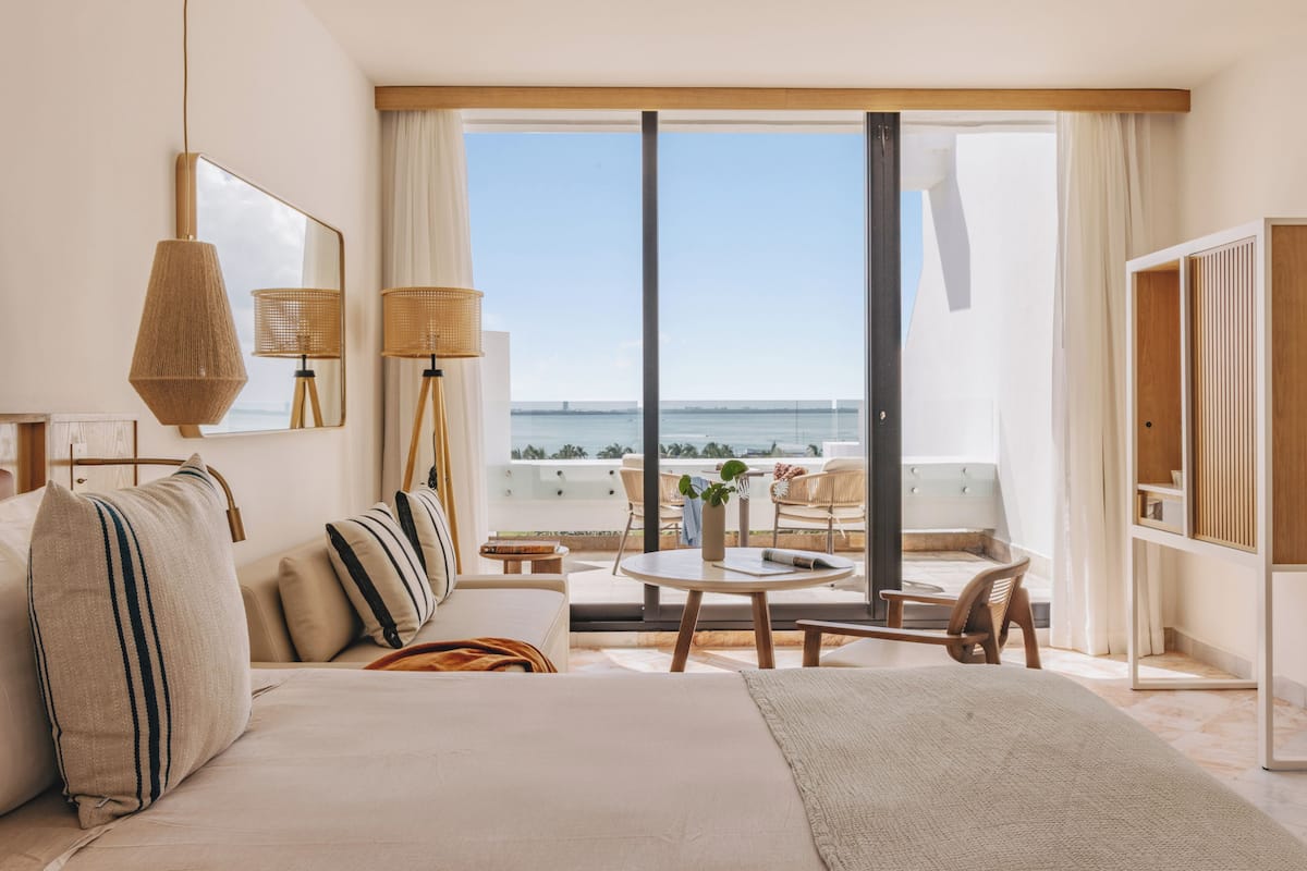 Quarto de hotel com uma cama, uma mesa redonda, uma cadeira, um sofá, uma janela grande com vista para o mar e uma varanda.