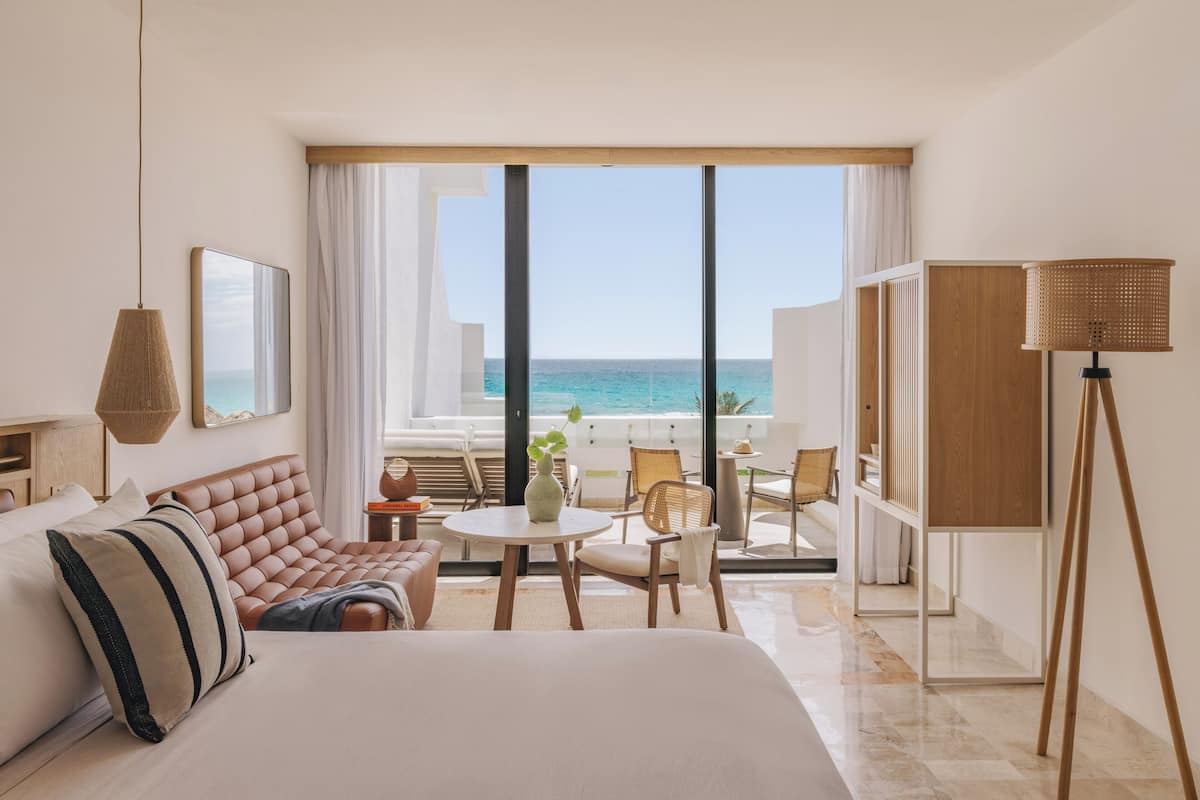 Quarto de hotel com cama, sofá, mesa de jantar com cadeiras, luminária e vista para o mar.