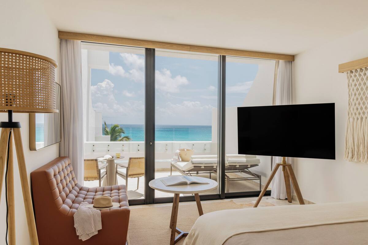 Quarto de hotel com uma janela grande, sofá, televisão e varanda com vista para o mar.