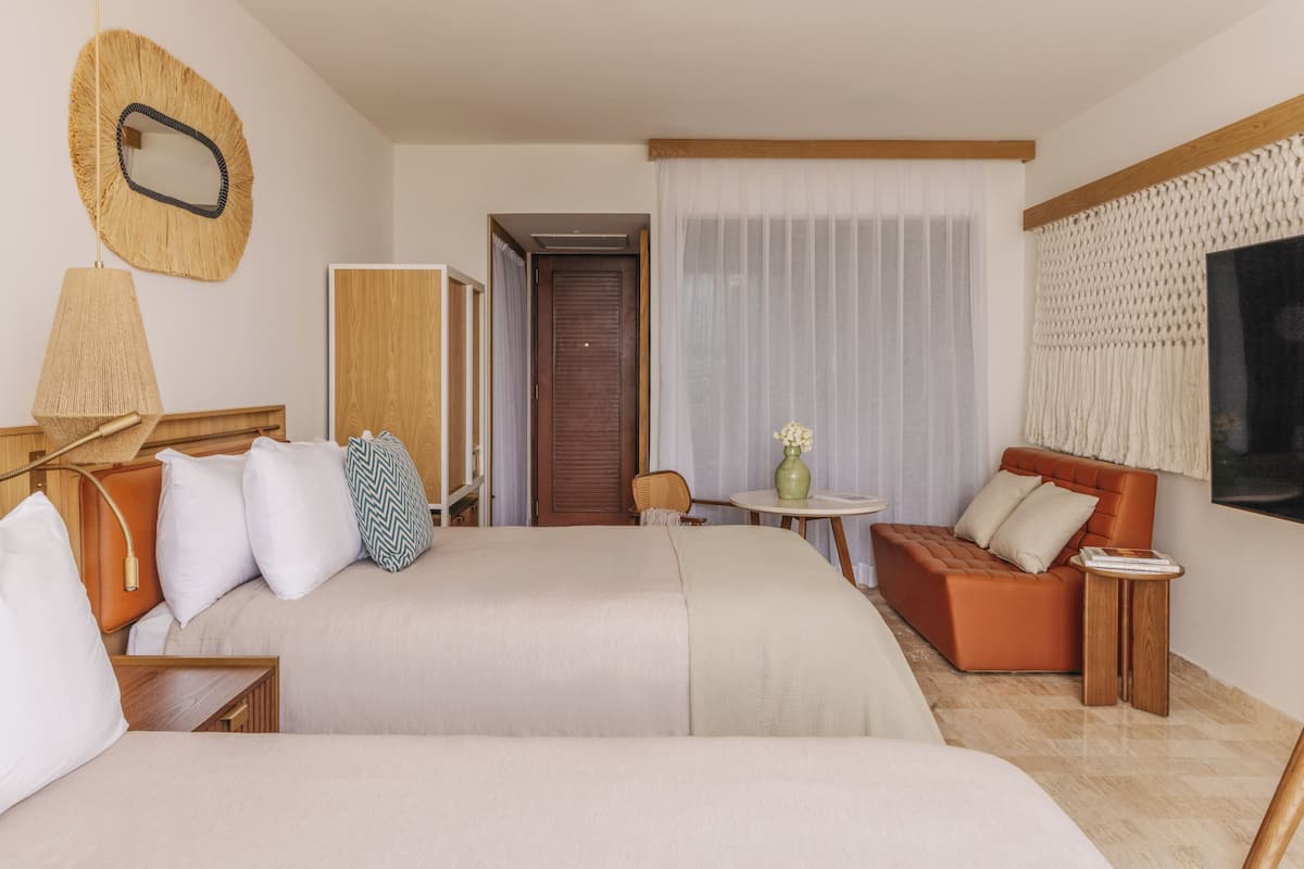 Quarto de hotel com duas camas, um sofá, uma mesa pequena, um abajur, um espelho e uma janela grande com cortinas.