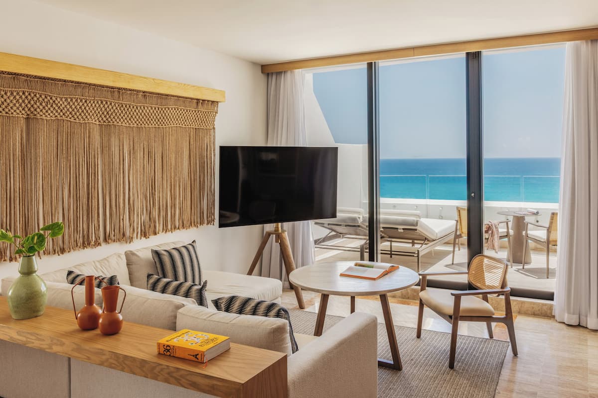 Quarto de hotel moderno com televisão grande, sofá, área de refeições e varanda com vista para o mar.