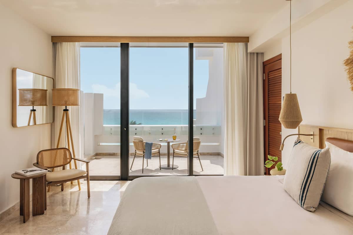 Quarto de hotel com cama, escrivaninha com cadeira, mesa com cadeiras e varanda com vista para o oceano.
