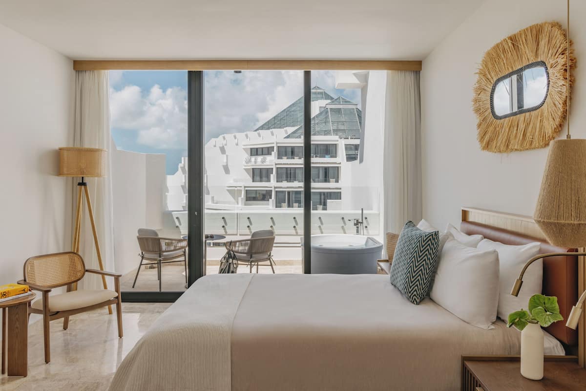 Quarto de hotel com uma cama grande, vista de um edifício através de uma porta de vidro, um espelho redondo e uma pequena mesa com abajur.