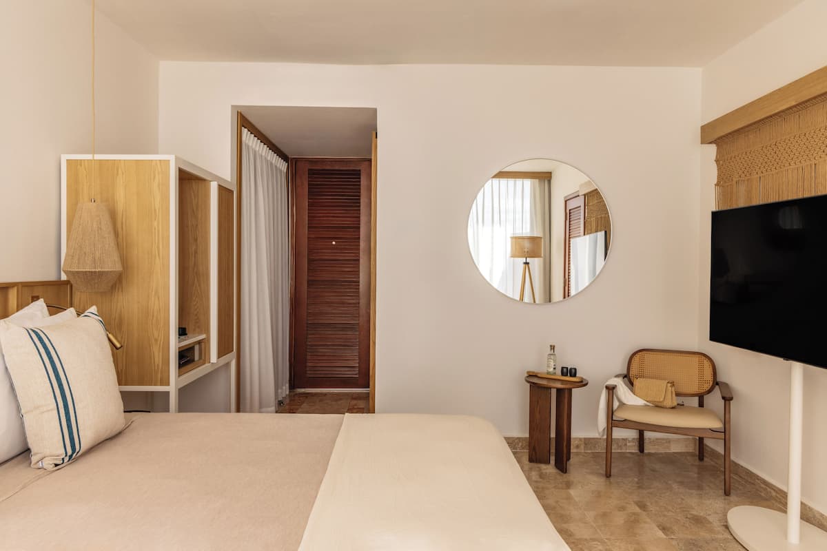 Quarto de hotel com cama, cadeira, televisão, espelho e guarda-roupa.