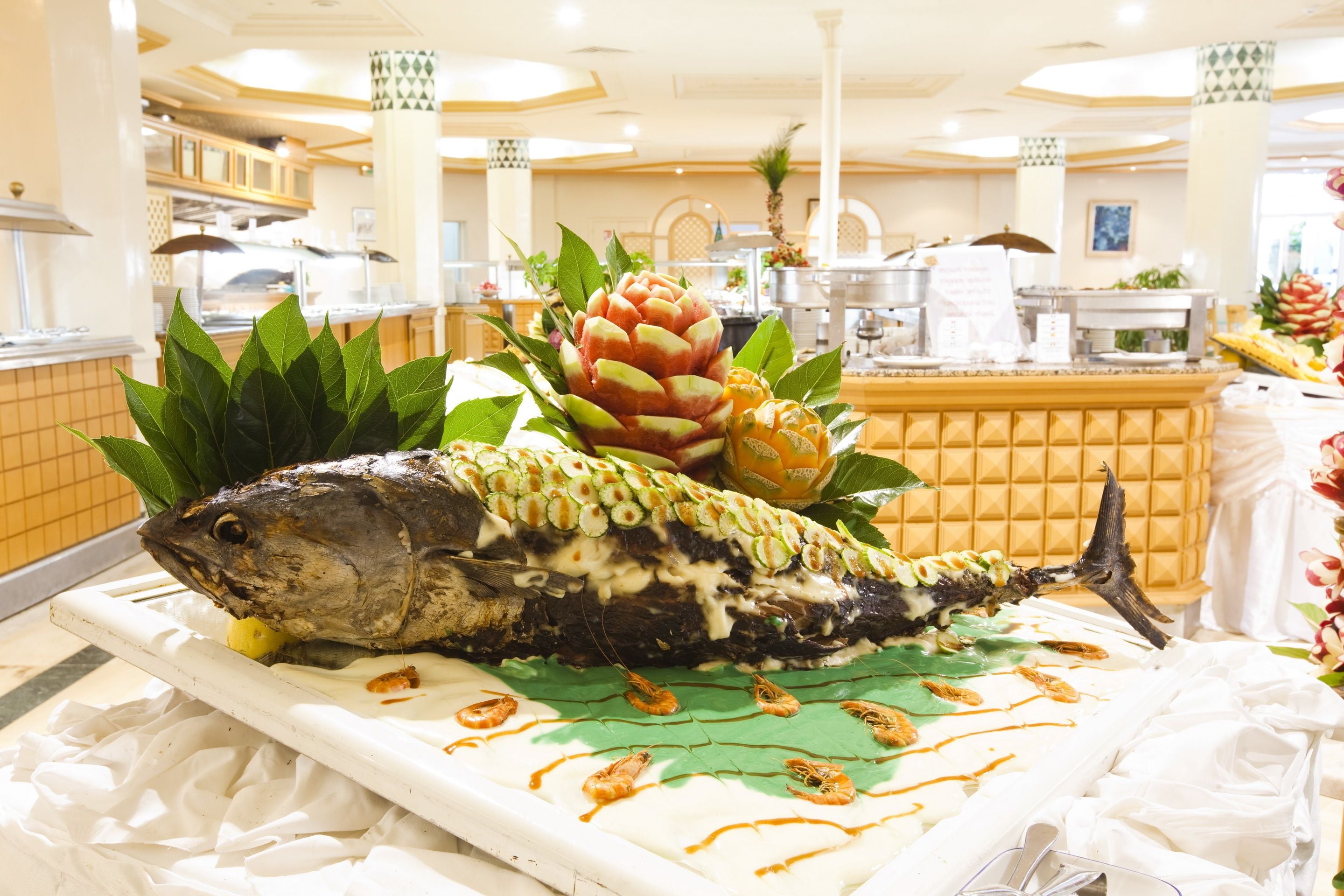 Prato de frutos do mar com peixe inteiro, camarão e abacaxi sobre um suporte branco.