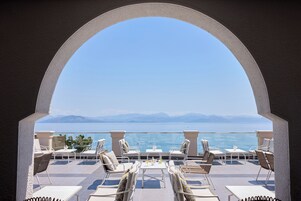 Terraço com vista para o mar e montanhas, mobiliado com cadeiras e mesas brancas.