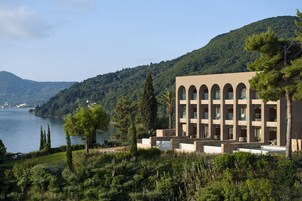 Um edifício moderno com várias varandas voltadas para um lago, cercado por vegetação exuberante e colinas.