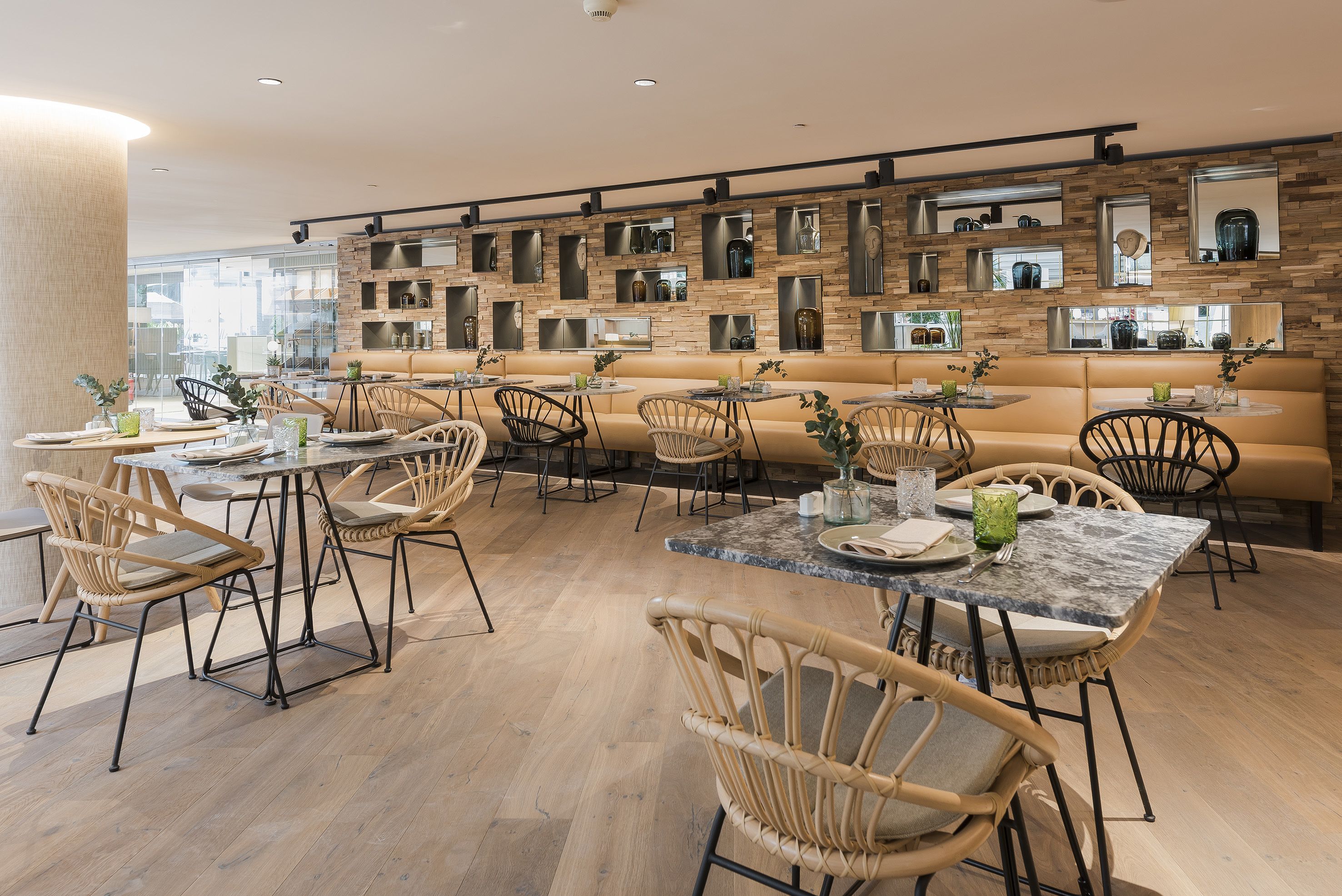 Restaurante moderno com cadeiras de vime, mesas de mármore e parede de tijolos com espelhos e prateleiras.