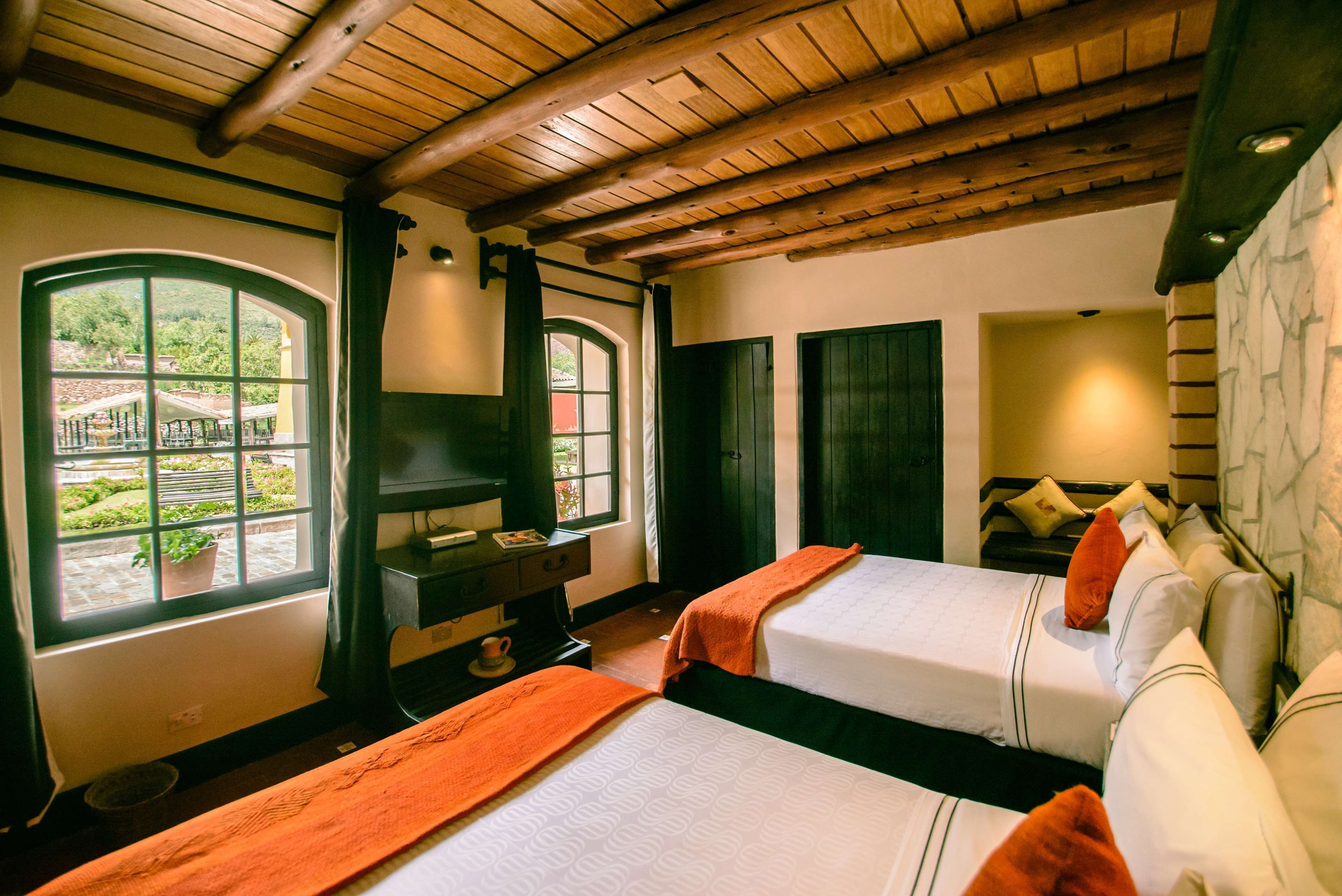 Quarto com duas camas, uma escrivaninha, janela com cortinas e vista para o verde lá fora.