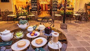 Um café ao ar livre com mesas preparadas para chá e café, rodeado por cadeiras e plantas em vasos.