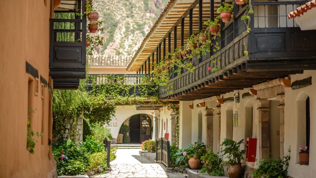 Uma passagem estreita com uma varanda de madeira decorada com plantas em vasos pendurados, que leva a uma entrada de edifício.