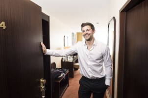 Quarto de hotel com porta de madeira marcada com o número 4, uma cama com uma mala, uma escrivaninha com abajur e um quadro na parede.