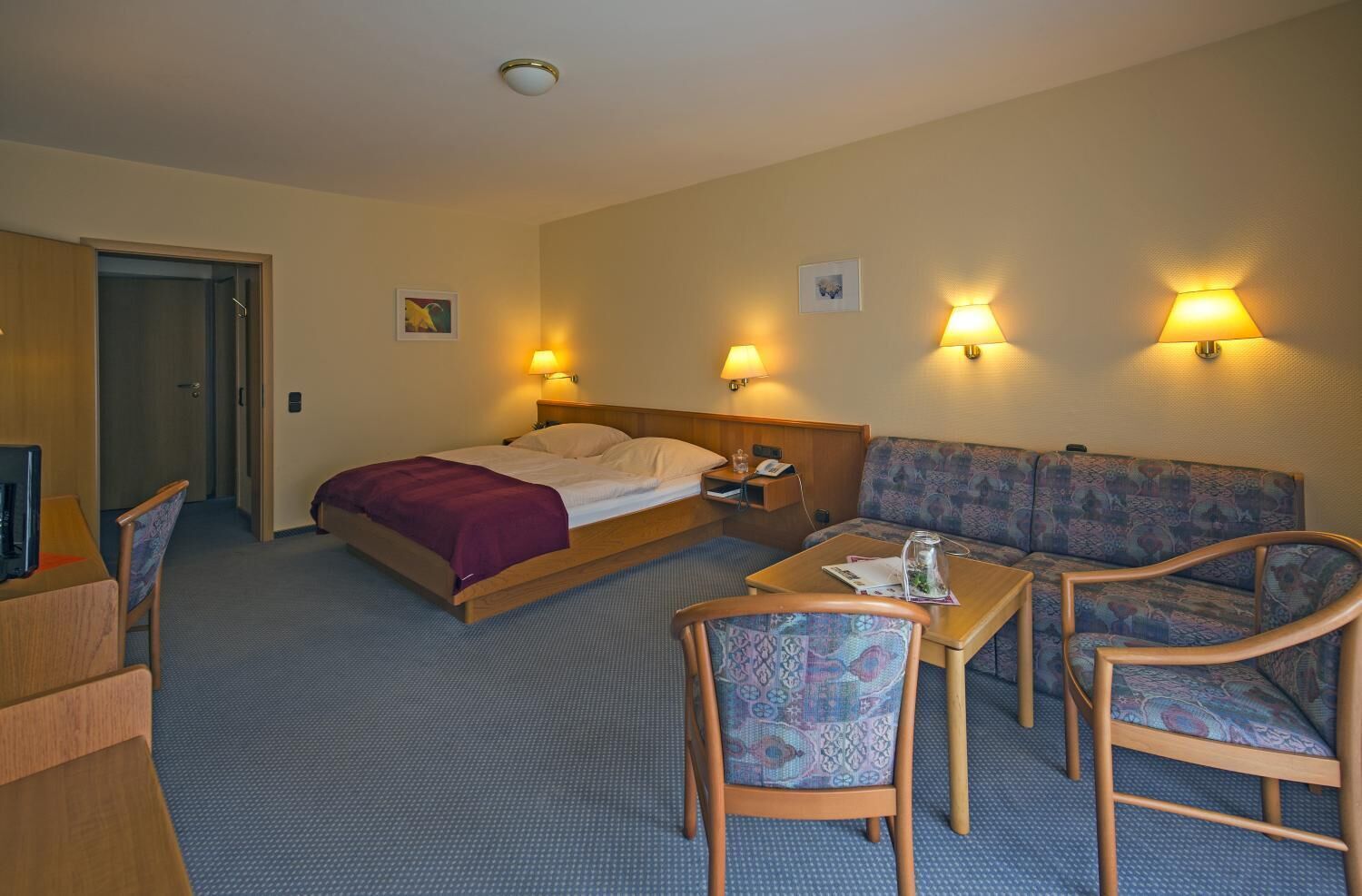 Quarto de hotel com uma cama, um sofá, duas poltronas, uma mesinha e uma escrivaninha com cadeira. Possui duas luminárias de parede e uma luminária no teto.