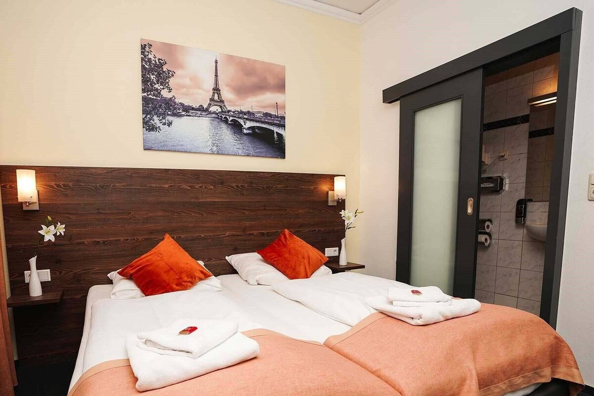 Quarto de hotel com uma cama, duas almofadas, uma mesa de cabeceira, um vaso com flores e um quadro da Torre Eiffel, além de uma porta de banheiro.