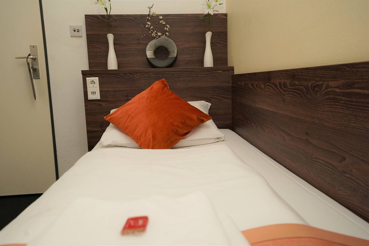 Cama bem arrumada com lençóis brancos e um travesseiro laranja, cabeceira de madeira e um pequeno vaso com flores na parede.