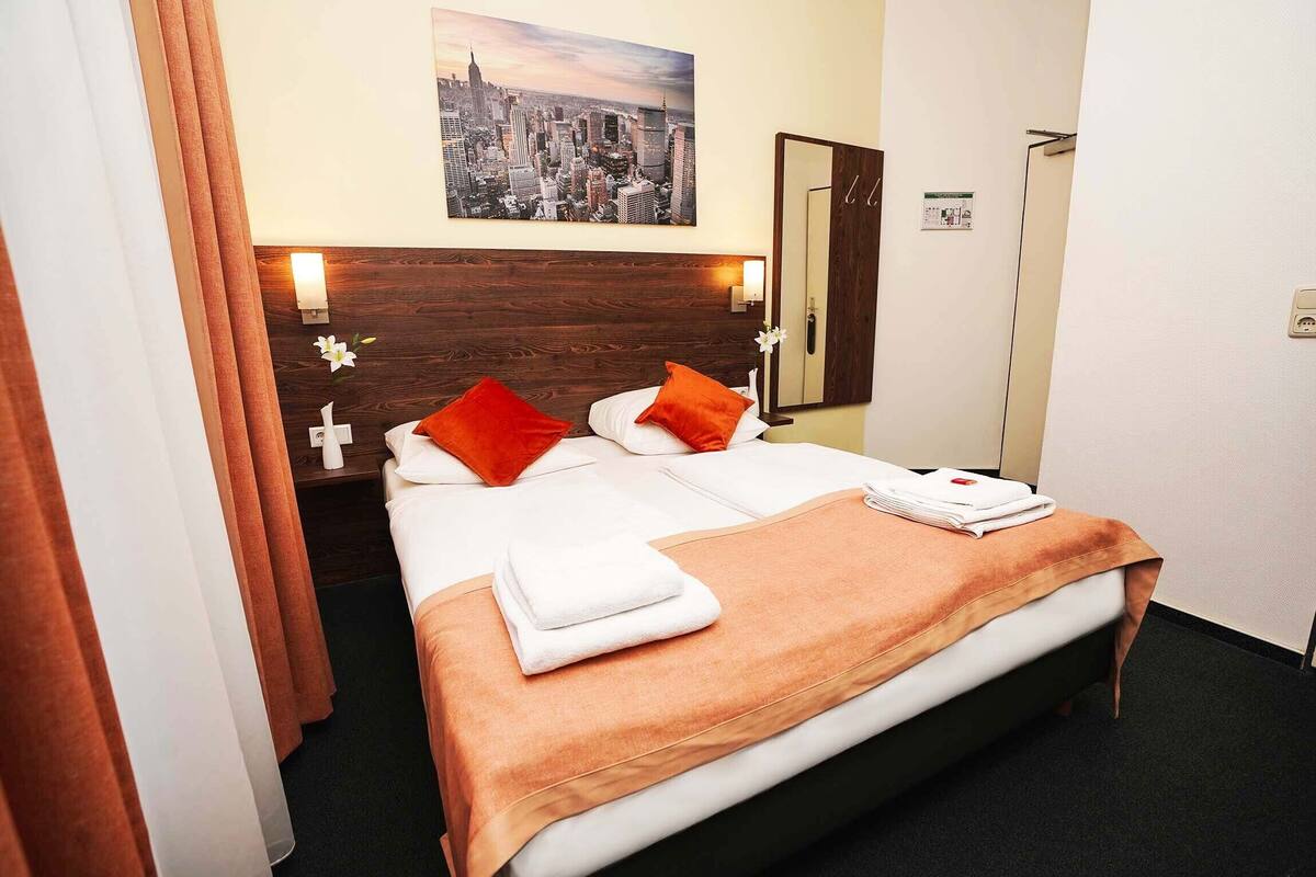 Quarto de hotel com uma cama grande, duas almofadas, mesa de cabeceira, espelho e uma pintura com vista para a cidade.