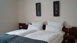 Quarto de hotel com duas camas de solteiro, uma cabeceira de madeira, mesa de cabeceira com abajur e telefone fixado na parede.