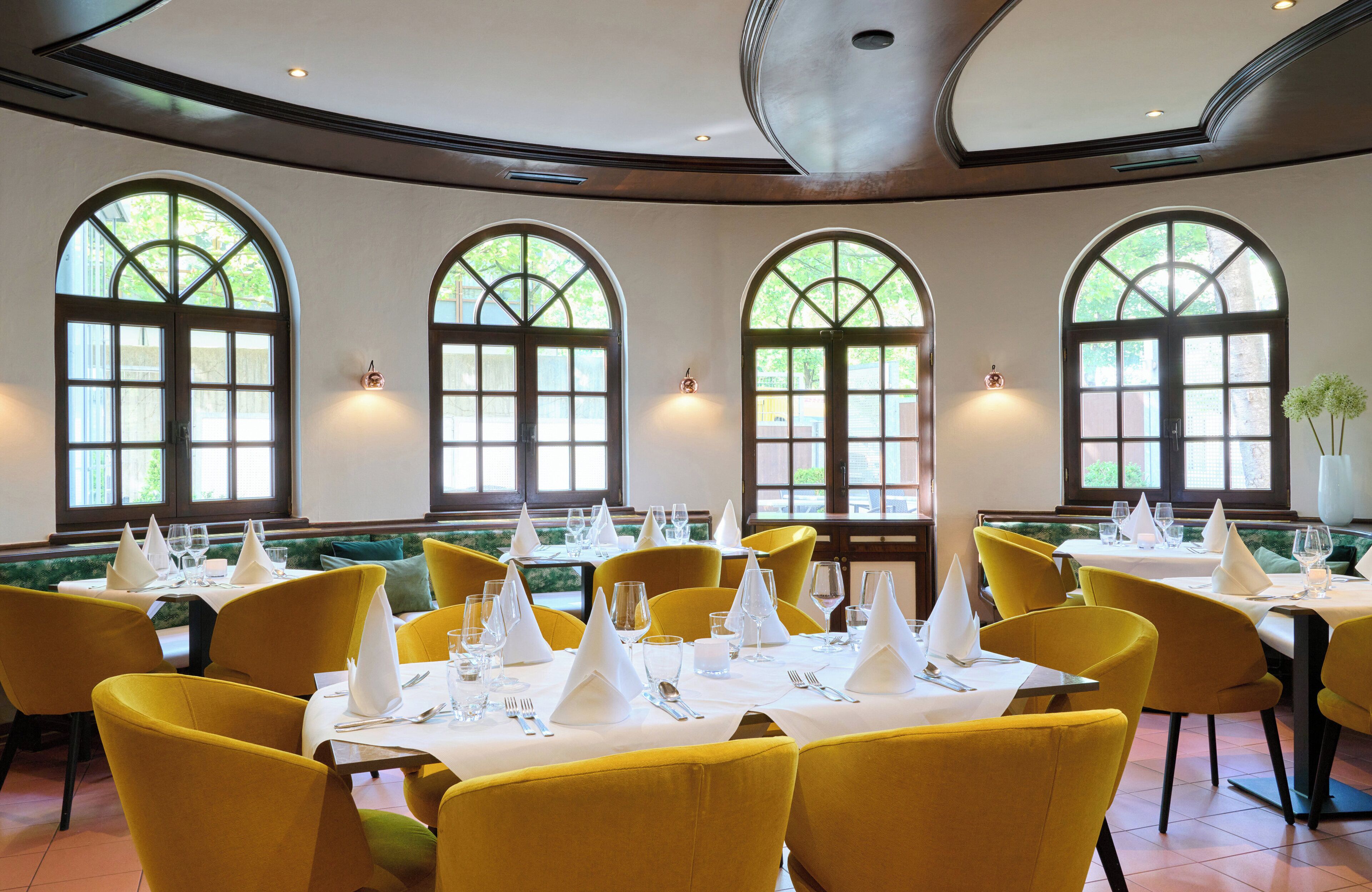 Uma sala de jantar com mesas preparadas para o serviço, cadeiras amarelas e amplas janelas em arco com luz natural.