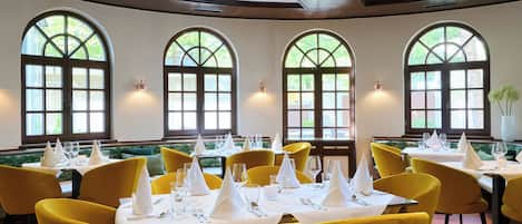 Uma sala de jantar com mesas preparadas para o serviço, cadeiras amarelas e amplas janelas em arco com luz natural.