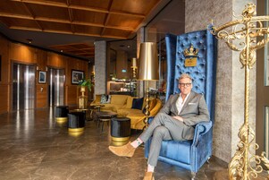 Um homem sentado em uma poltrona azul de veludo com emblema de coroa, em um saguão com lambris de madeira, um sofá amarelo e um abajur dourado.