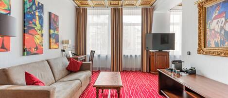 Quarto de hotel com carpete vermelho, sofá bege, mesa de madeira, televisão e obras de arte nas paredes.