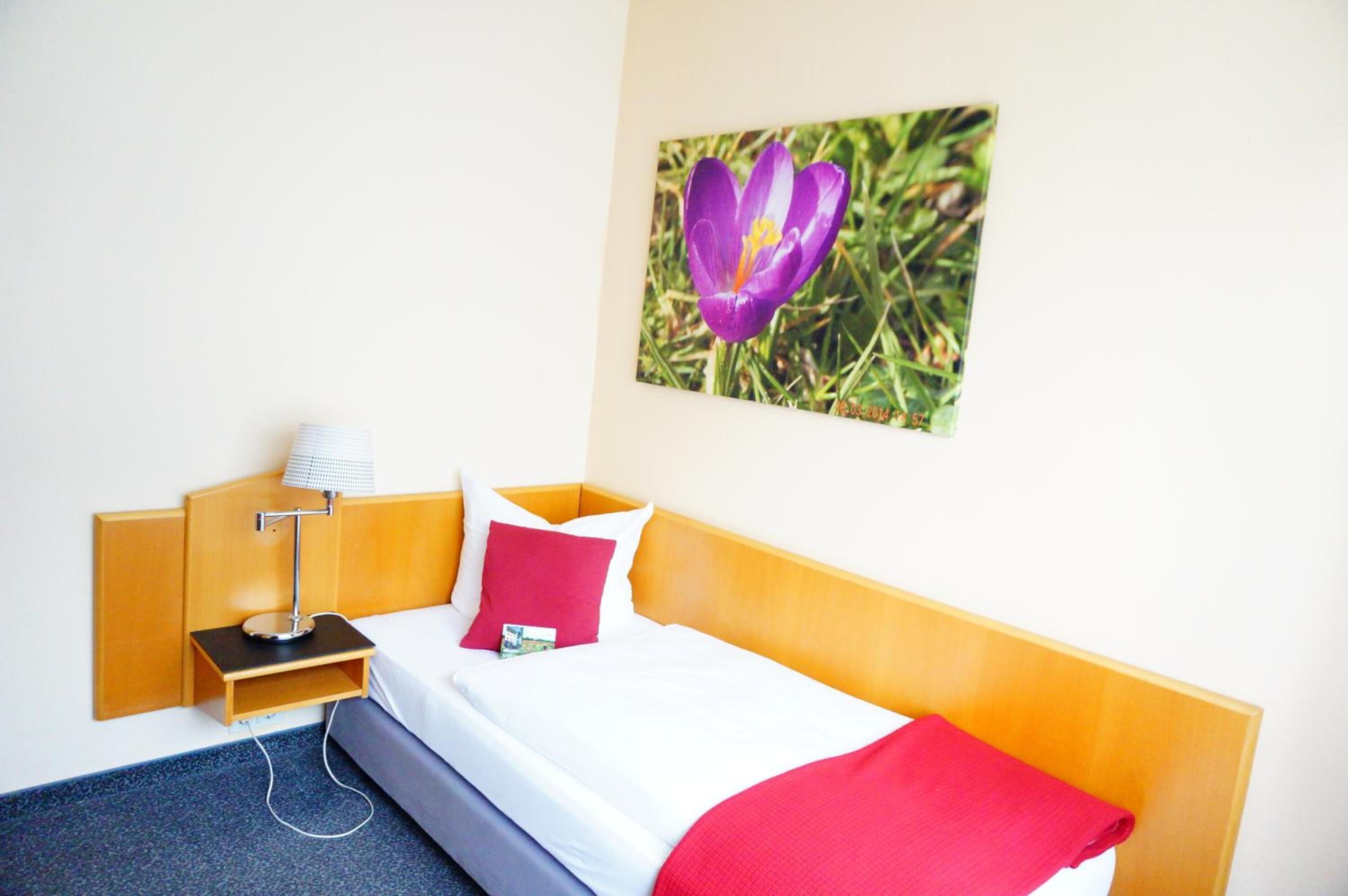 Quarto de hotel com uma cama, mesa de cabeceira, abajur e um quadro de uma flor roxa na parede.