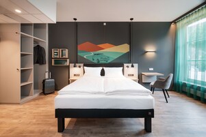 Quarto de hotel moderno com uma cama grande, uma escrivaninha e uma cadeira. Possui um guarda-roupa com prateleiras e espaço para pendurar roupas, e uma mala está visível.