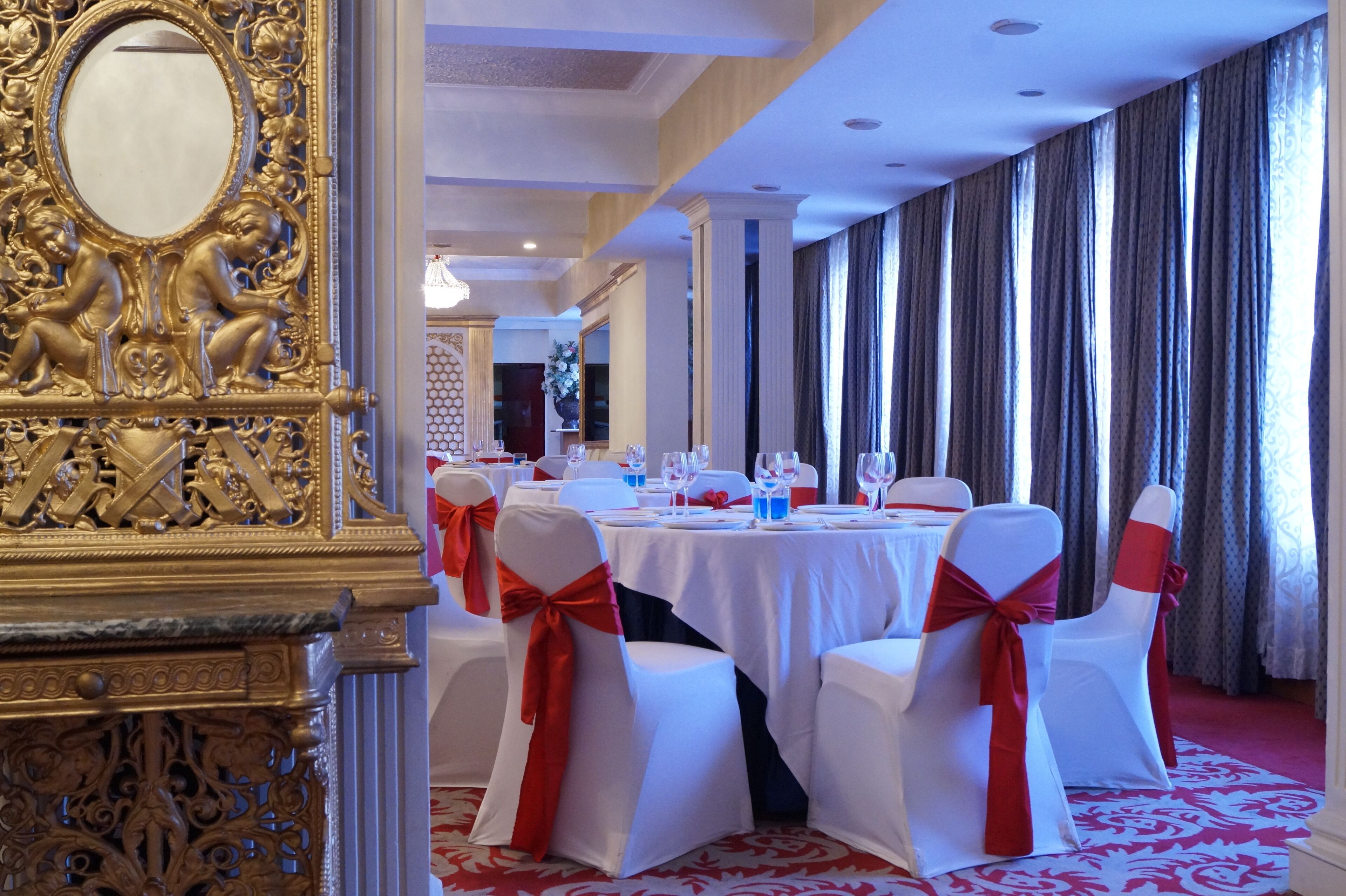 Uma sala de banquetes com mesas e cadeiras brancas, fitas vermelhas e um espelho dourado decorativo.