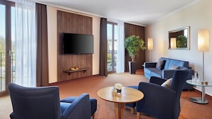 Quarto de hotel com televisão de tela plana na parede, um sofá, duas poltronas, uma mesinha redonda com um vaso e uma planta em vaso.
