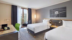 Quarto de hotel com duas camas, uma escrivaninha com luminária, uma cadeira e vista para árvores pela janela.